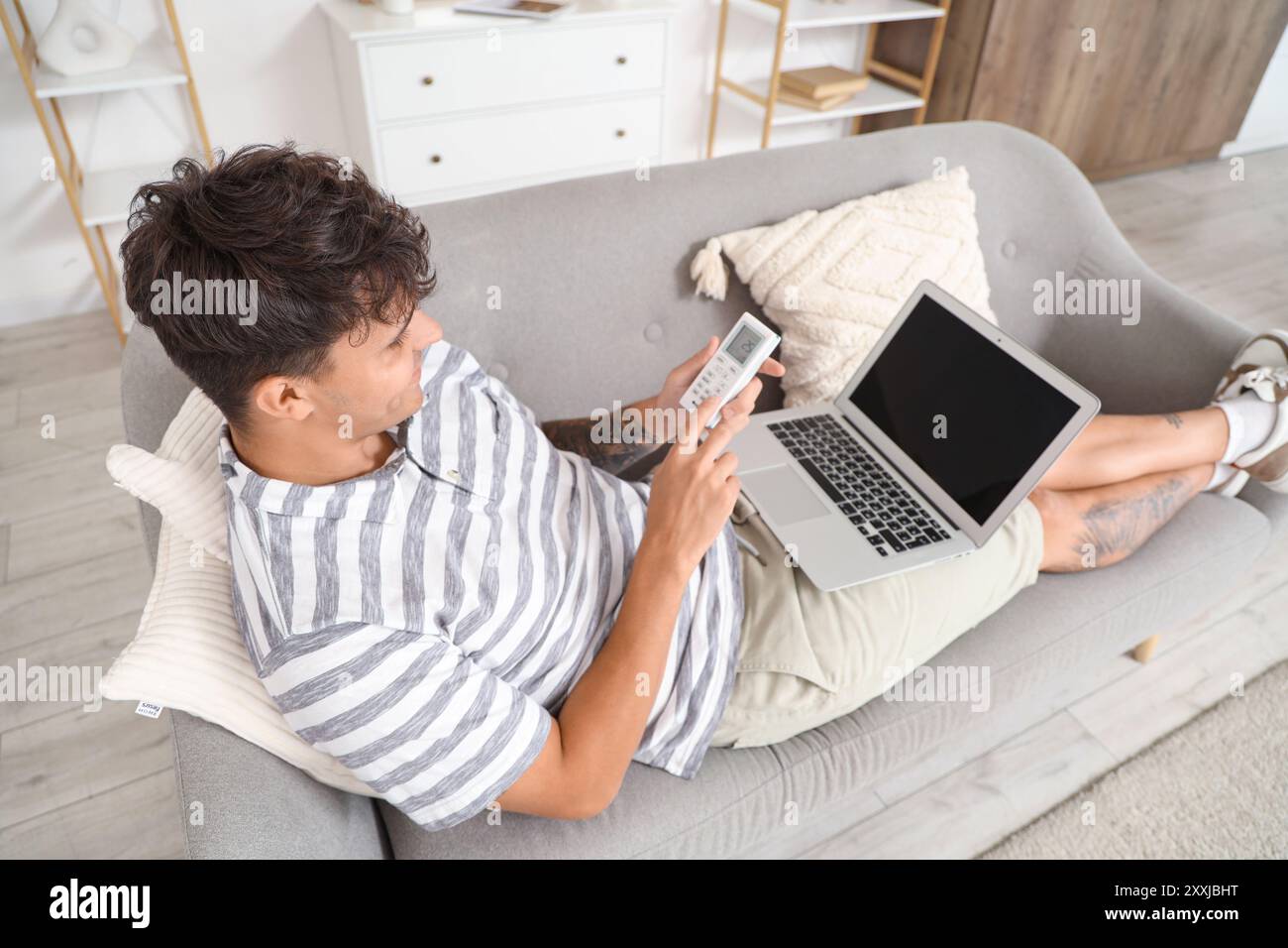Handsome young man with air conditioner remote control and laptop lying ...