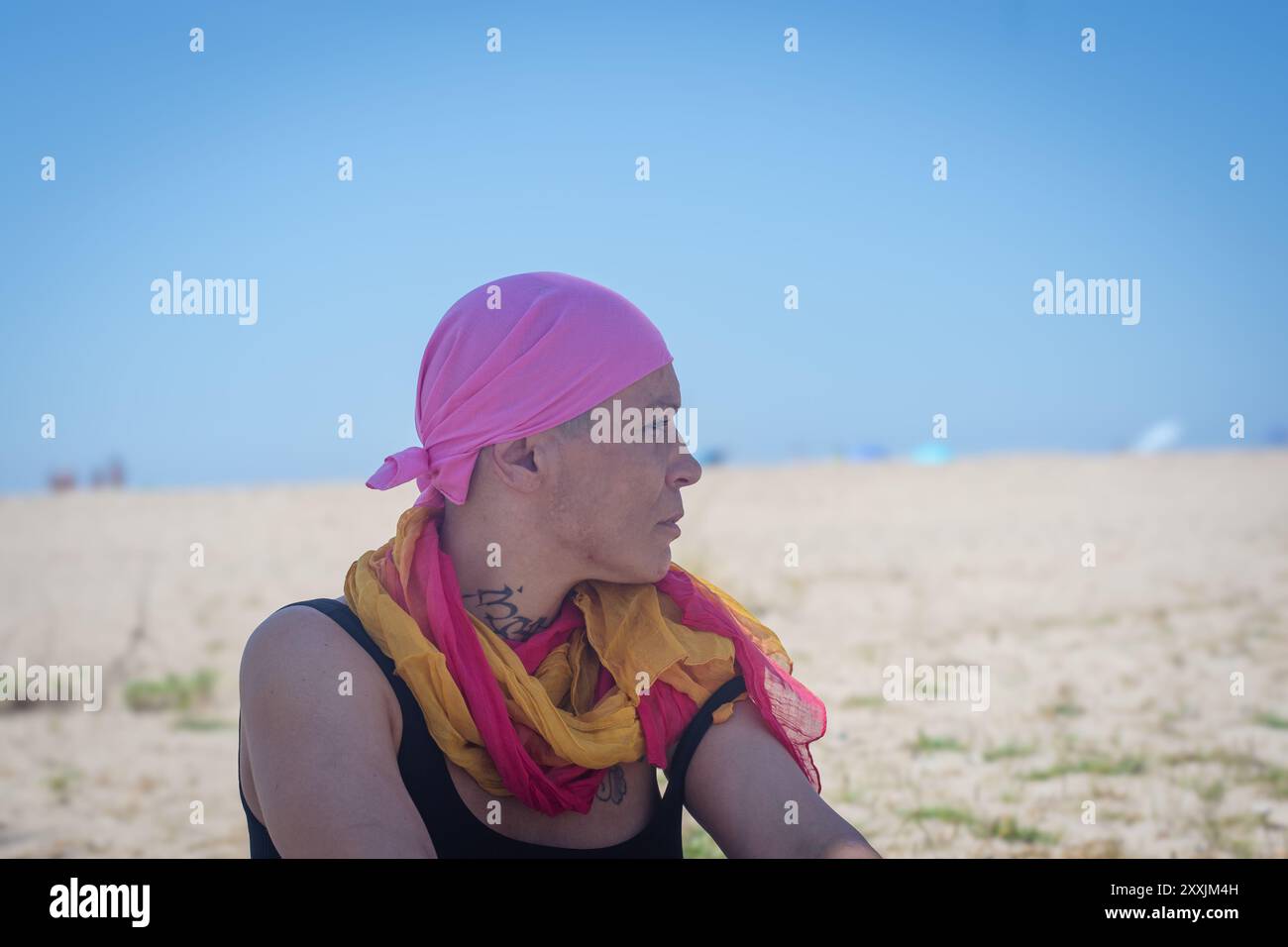 Cancer survivor in a pink headscarf sits quietly on the beach, gazing ...
