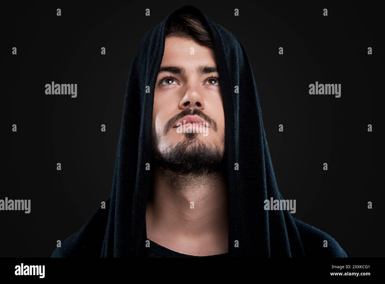 Mysterious man. Handsome young man in black hood looking up while standing against black ...