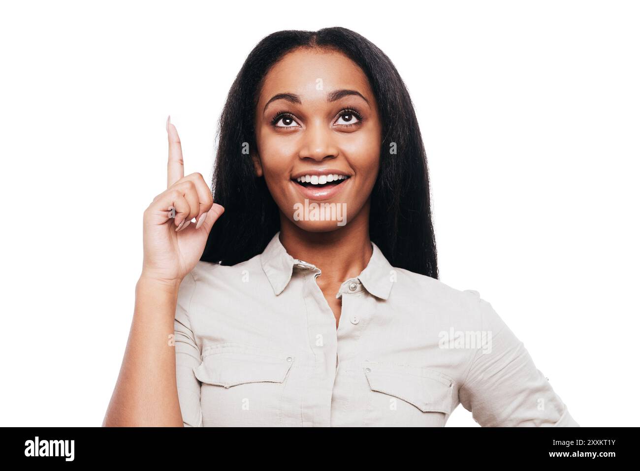 Just inspired. Happy young African woman pointing up and smiling while standing against white ...