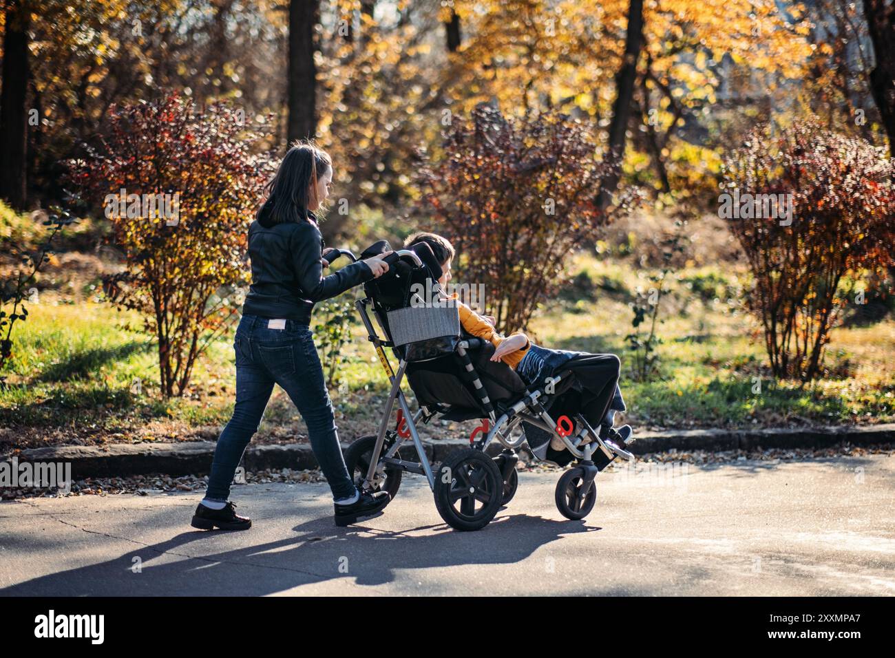 Sibling pushing child in wheelchair through park, accessible recreational spaces, mobility ...