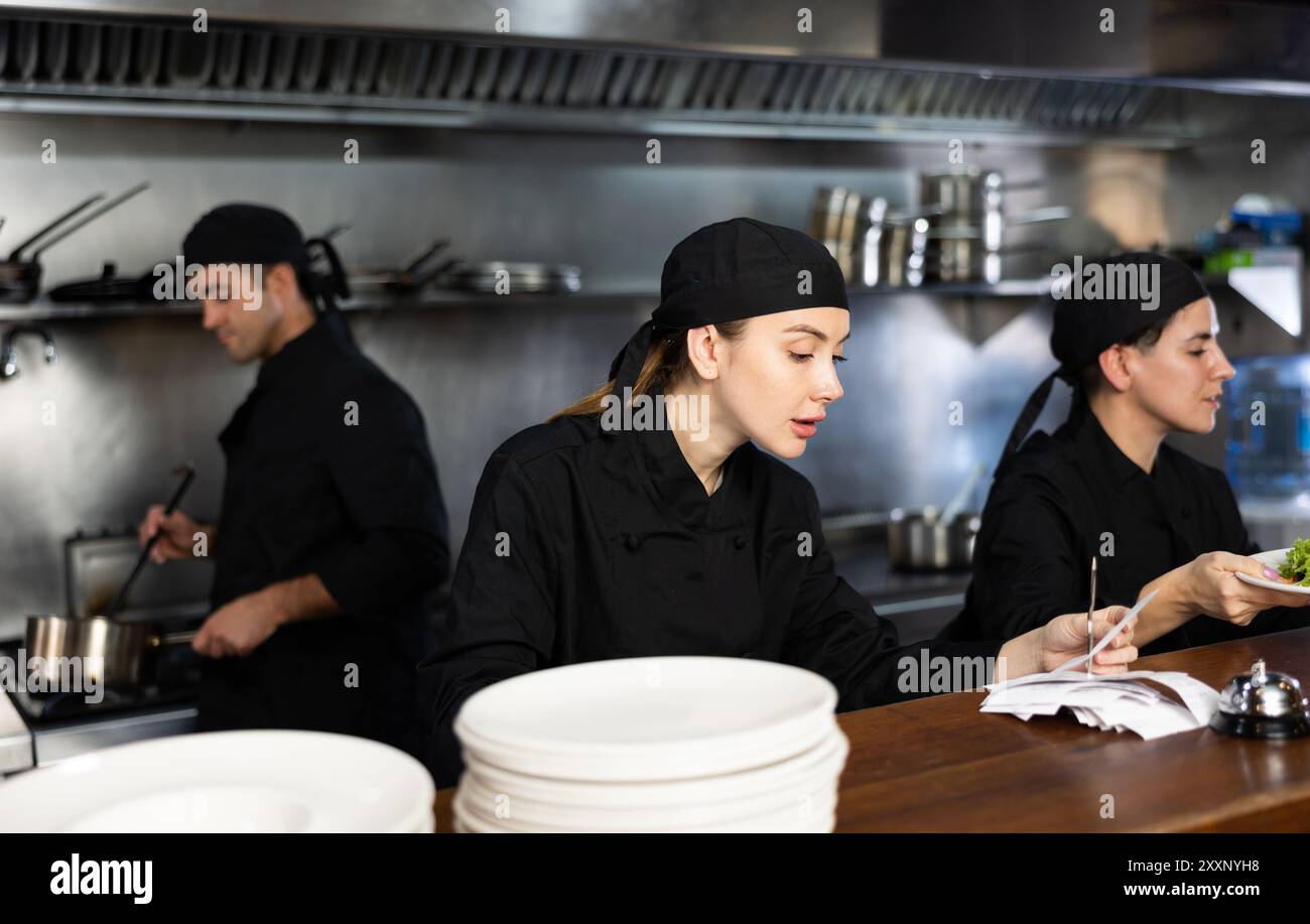 Female chef checks the prepared dish with the paid check in kitchen of ...