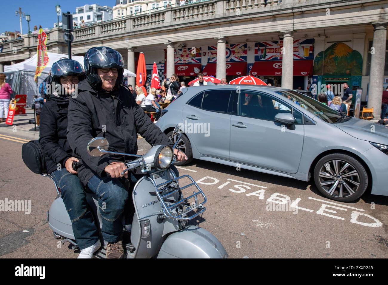 Two Mods riding a grey scooter ride past a grey car. Thousands attend ...