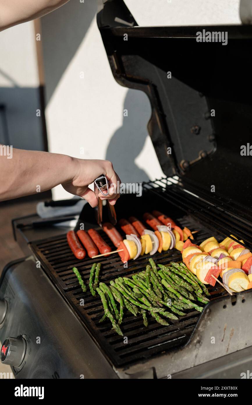 Hand using tongs to grill veggie skewers, and hot dogs on a barbecue ...