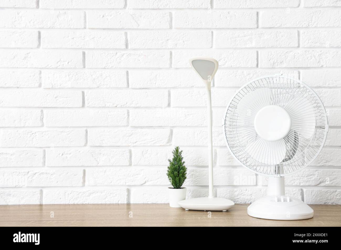 Electric fan on table with lamp and houseplant near white brick wall in ...