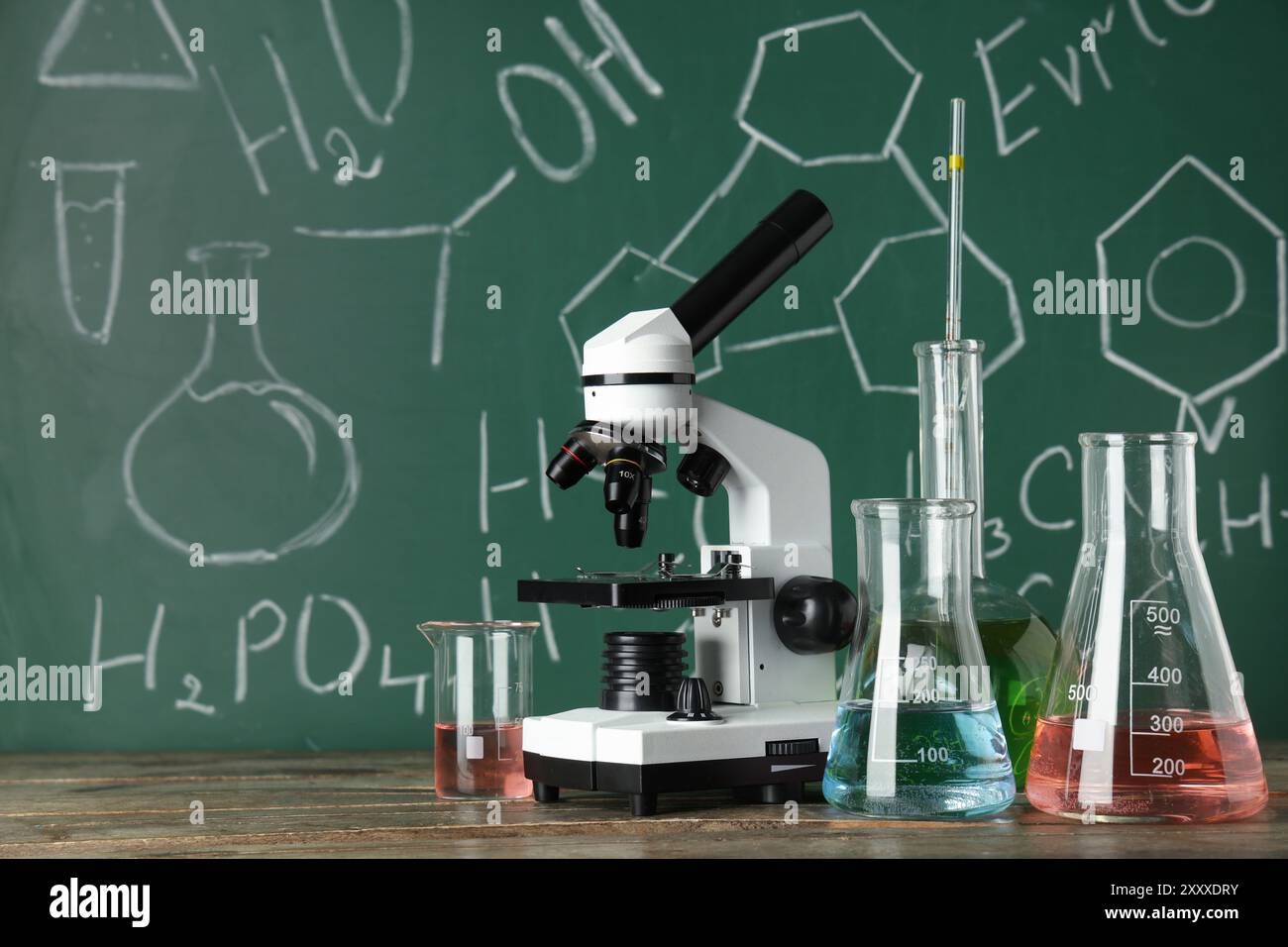 Modern microscope and flasks on wooden table near chalkboard with ...