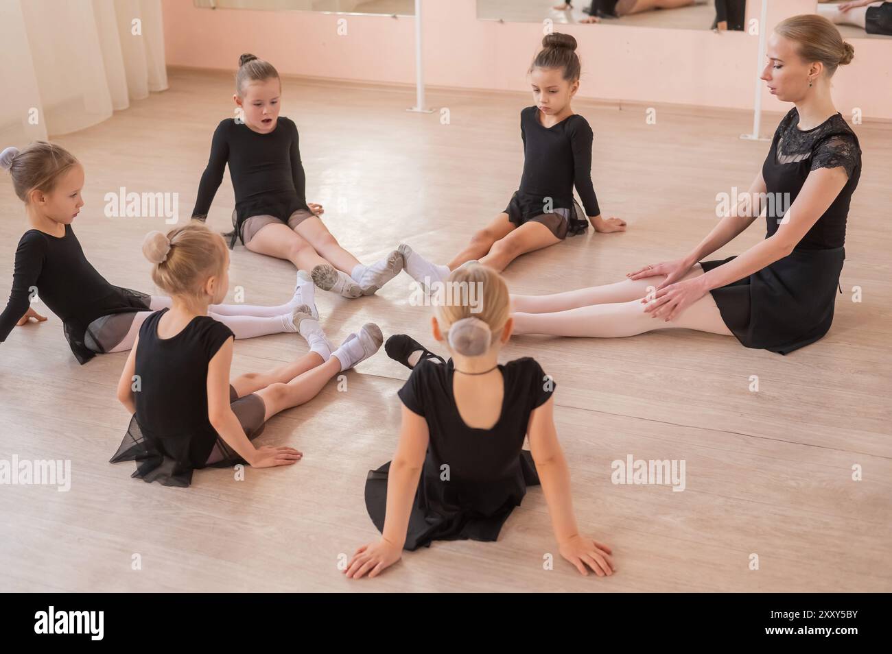 Caucasian woman and five little girls sit in a circle and do stretching ...