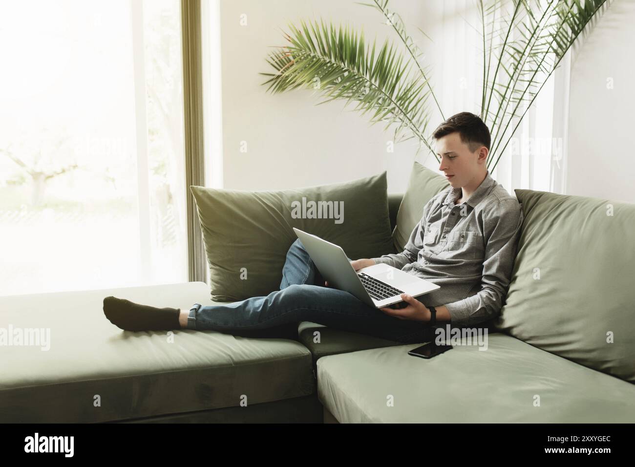 Young guy sitting at green sofa with gray laptop, black phone. Man is ...