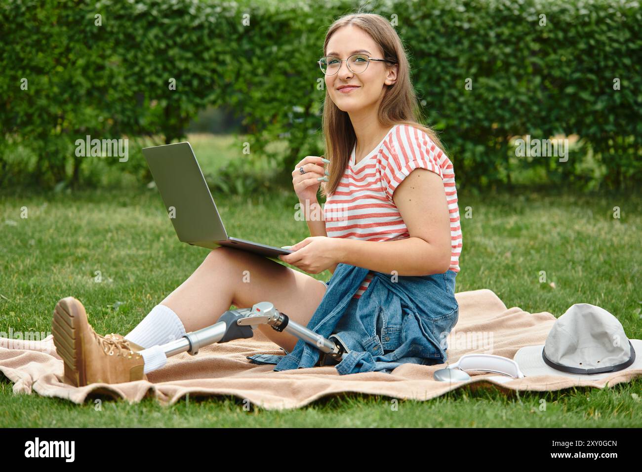 A young woman with a prosthetic leg sits on a blanket in a park, using ...
