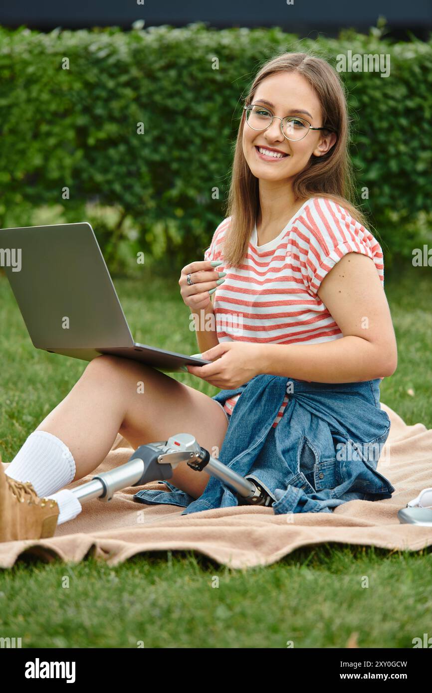 A young woman with a prosthetic leg sits on a blanket in a park, using ...