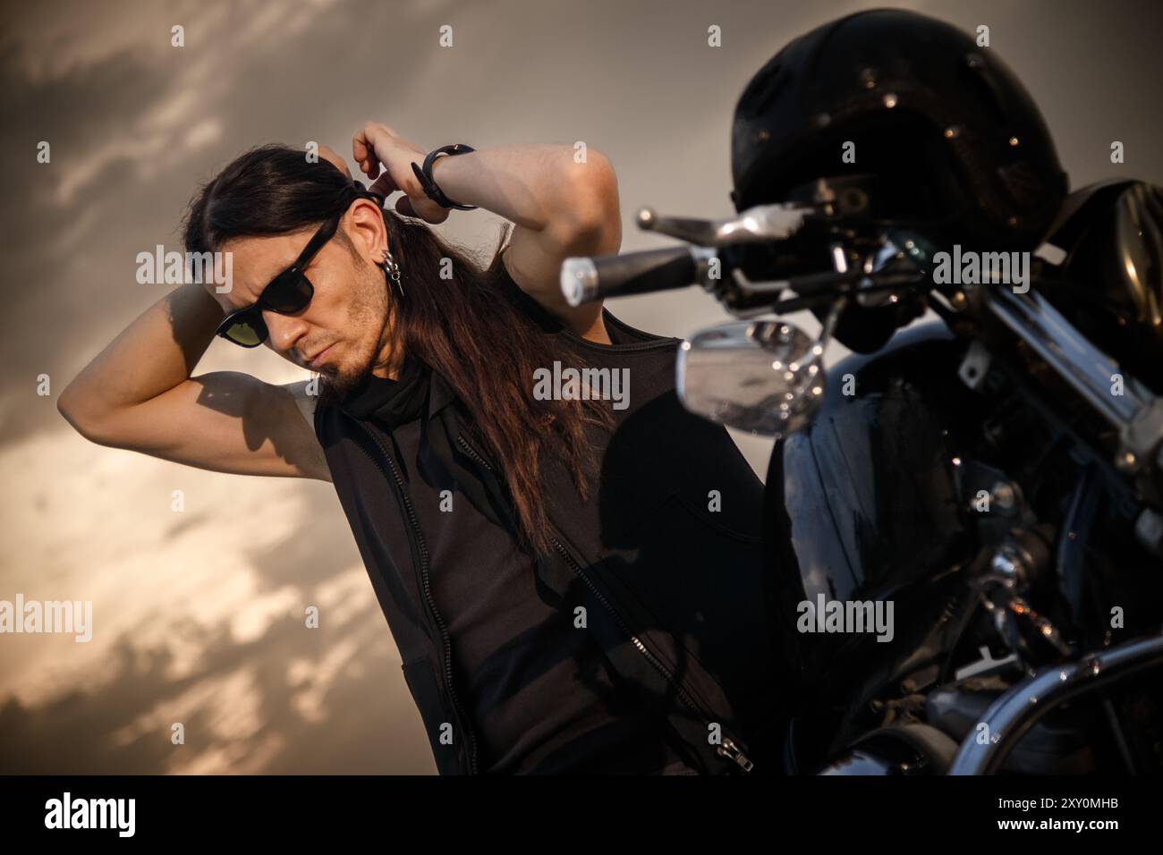 A biker with long hair leaning on his chopper-style motorcycle by the ...