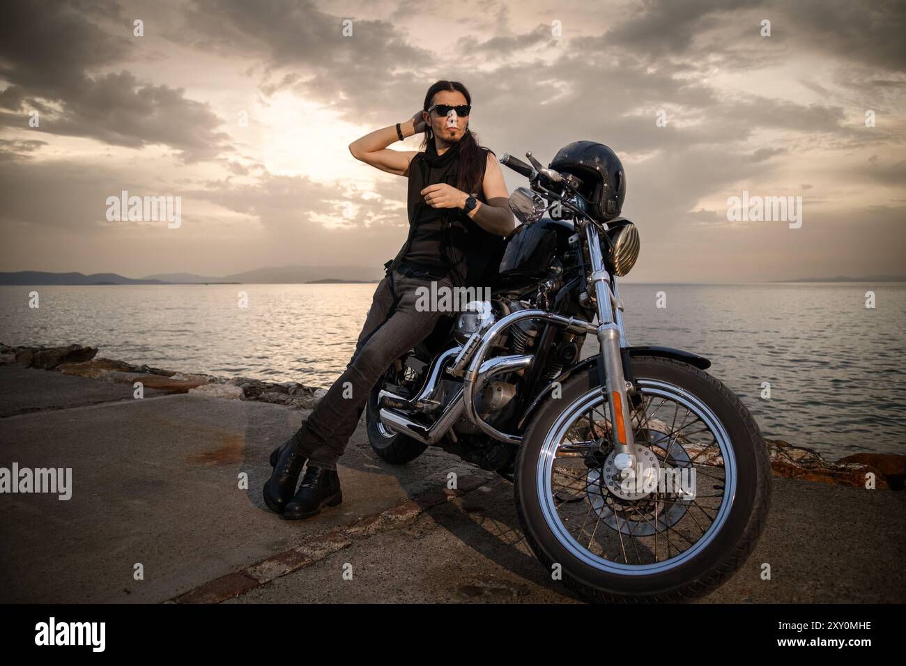 A biker with long hair leaning on his chopper-style motorcycle by the ...