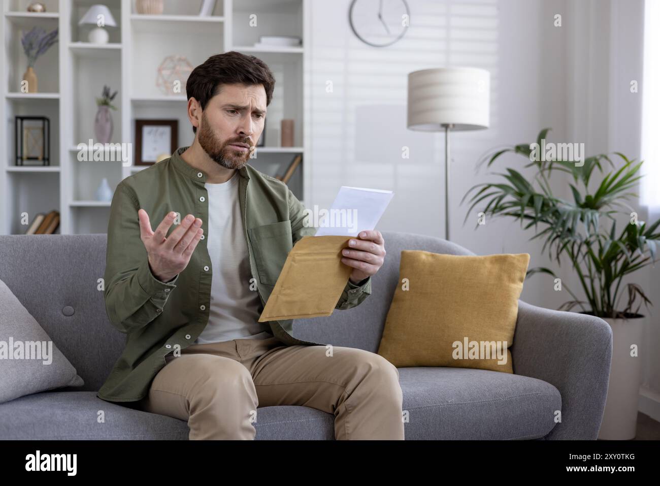 Man with beard sitting on couch looking at letter with confused ...