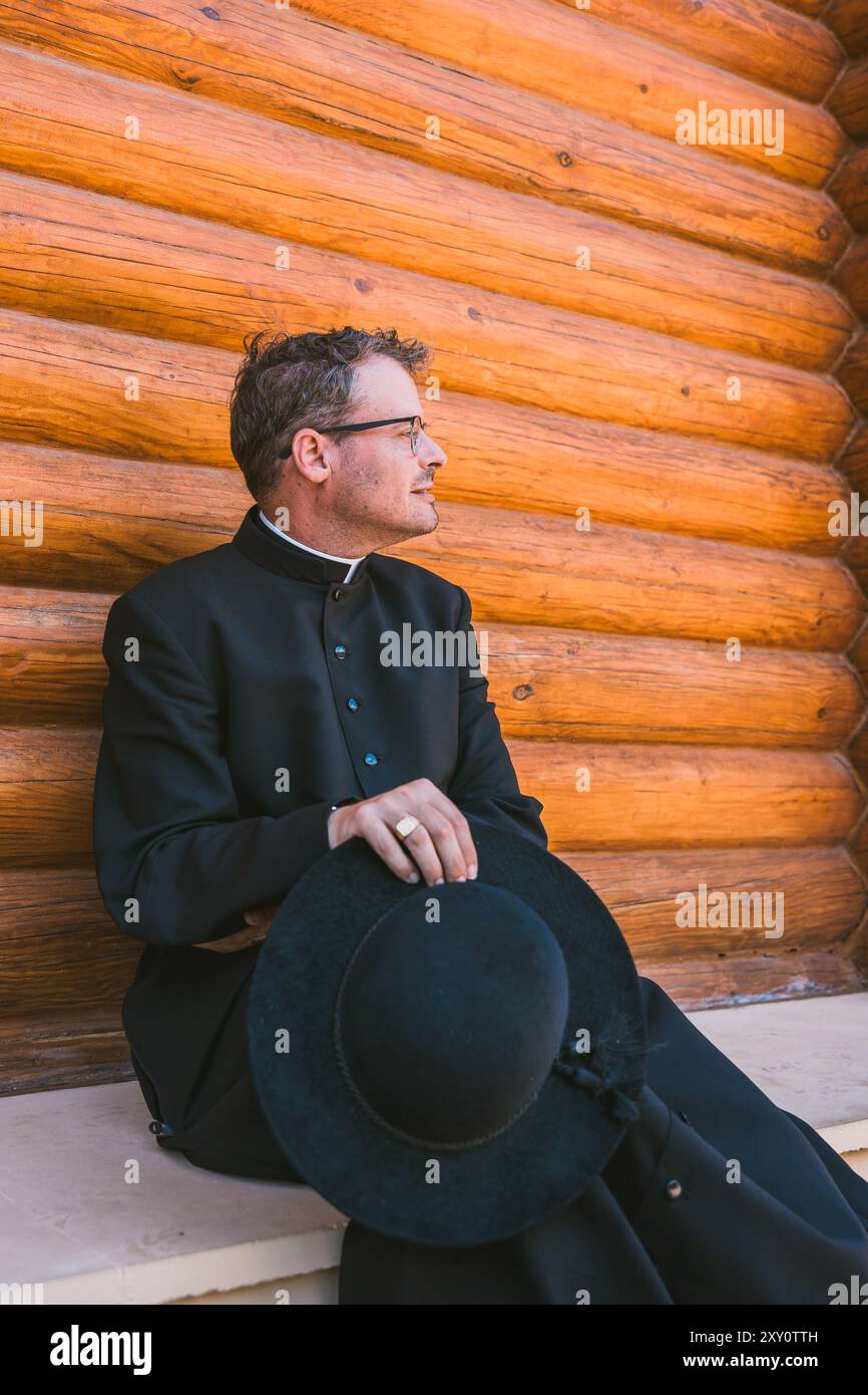 Catholic priest in contemplative pose, seated outside against a rustic ...