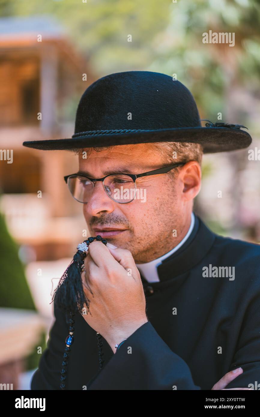 Catholic priest in a black cassock and hat, praying with a rosary ...
