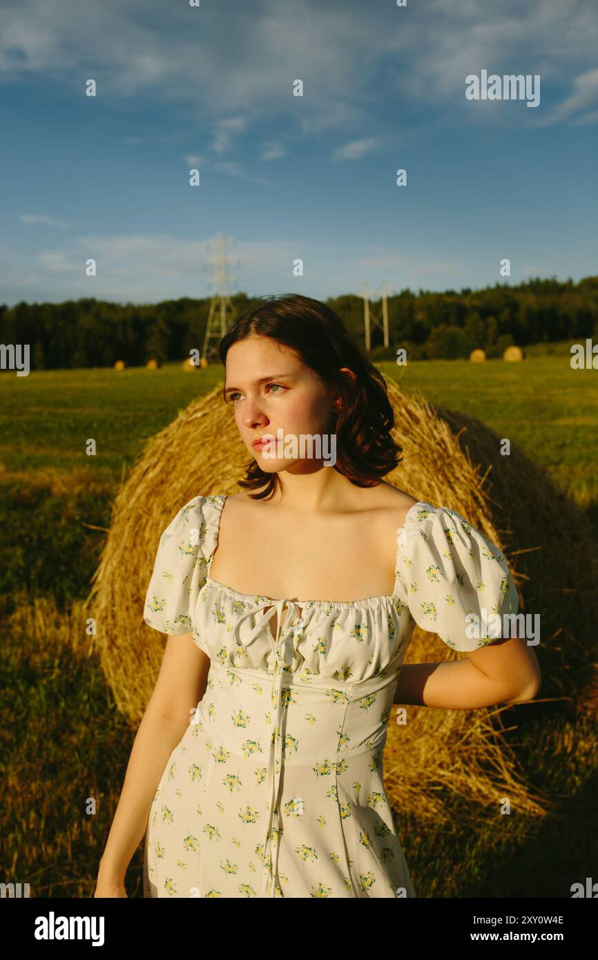 A young woman in a light floral prom dress stands beside a hay bale in ...