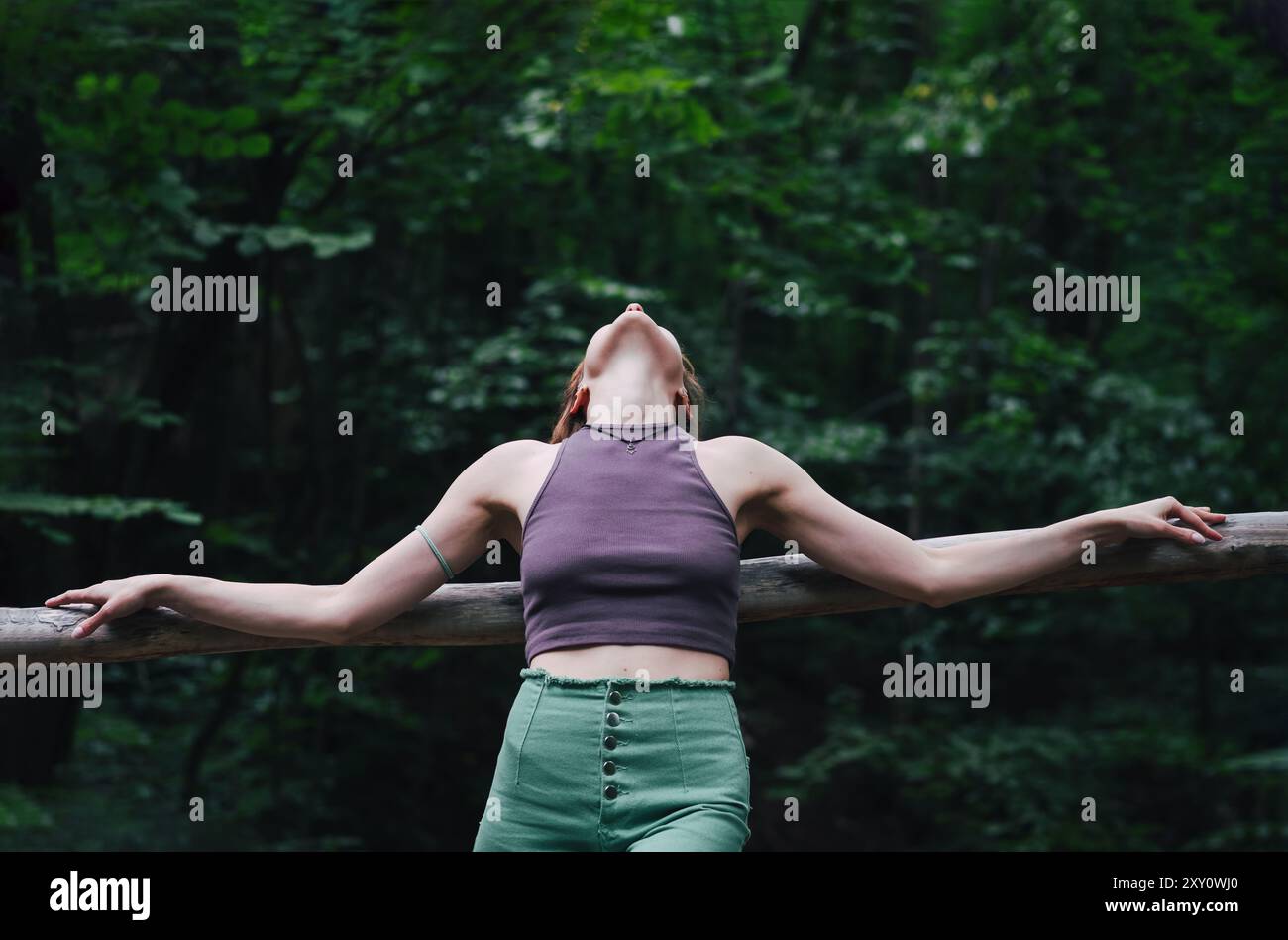 Unrecognizable woman on a wooden rail in a dense forest, head tilted ...