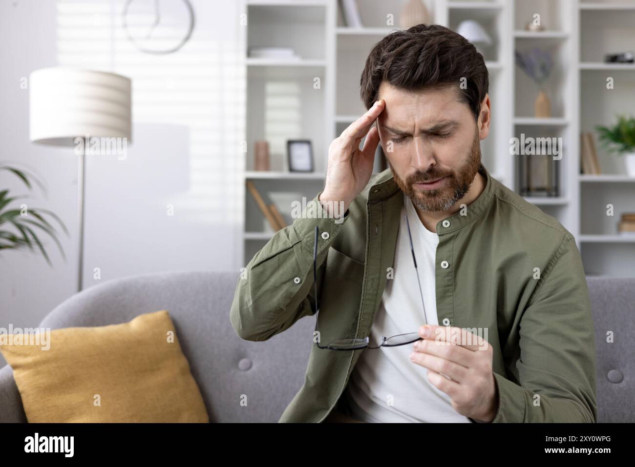 Man sitting on gray couch experiencing headache, holding glasses with ...