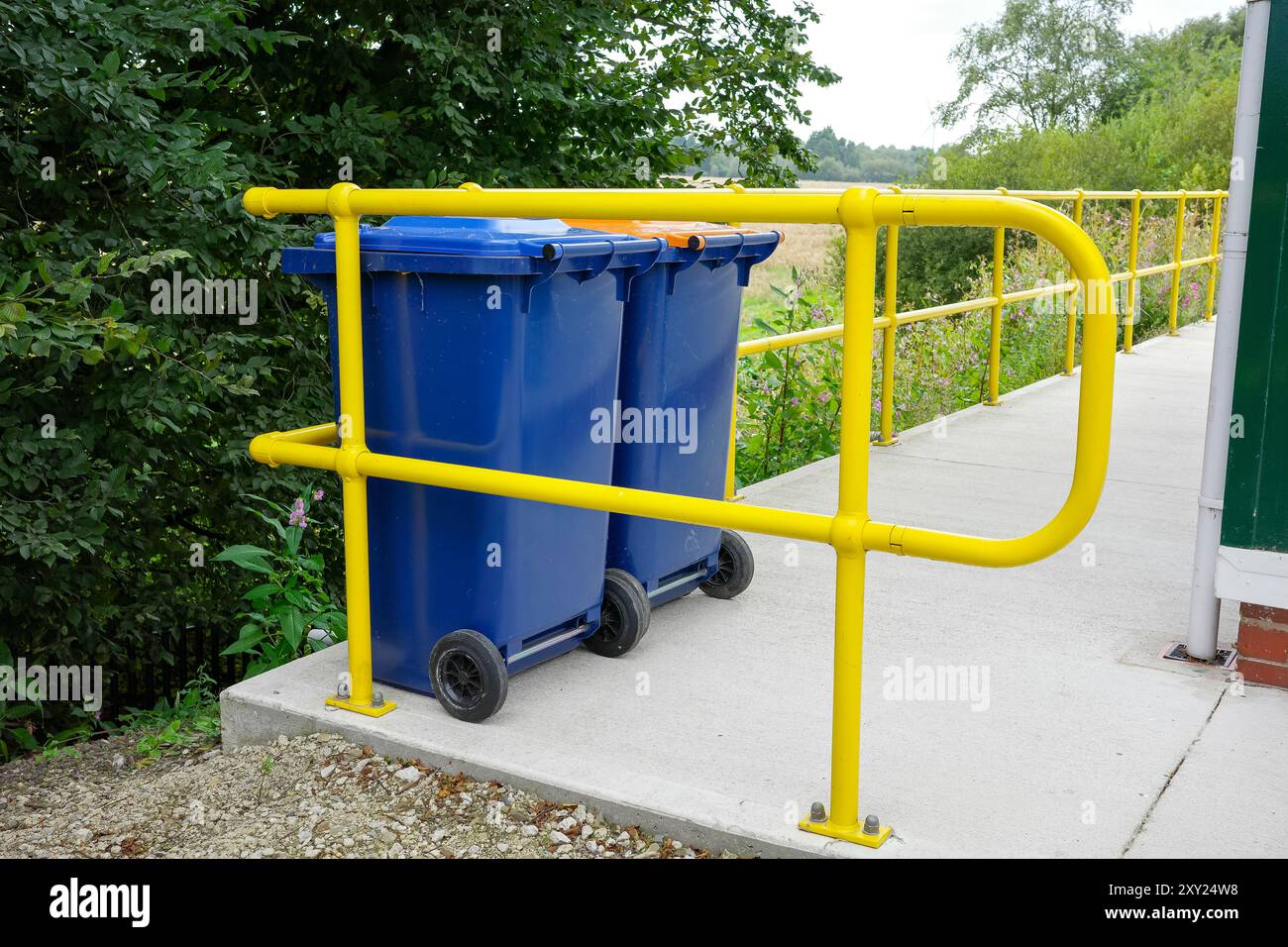 Two blue recycling bins placed next to yellow railing on a paved area ...