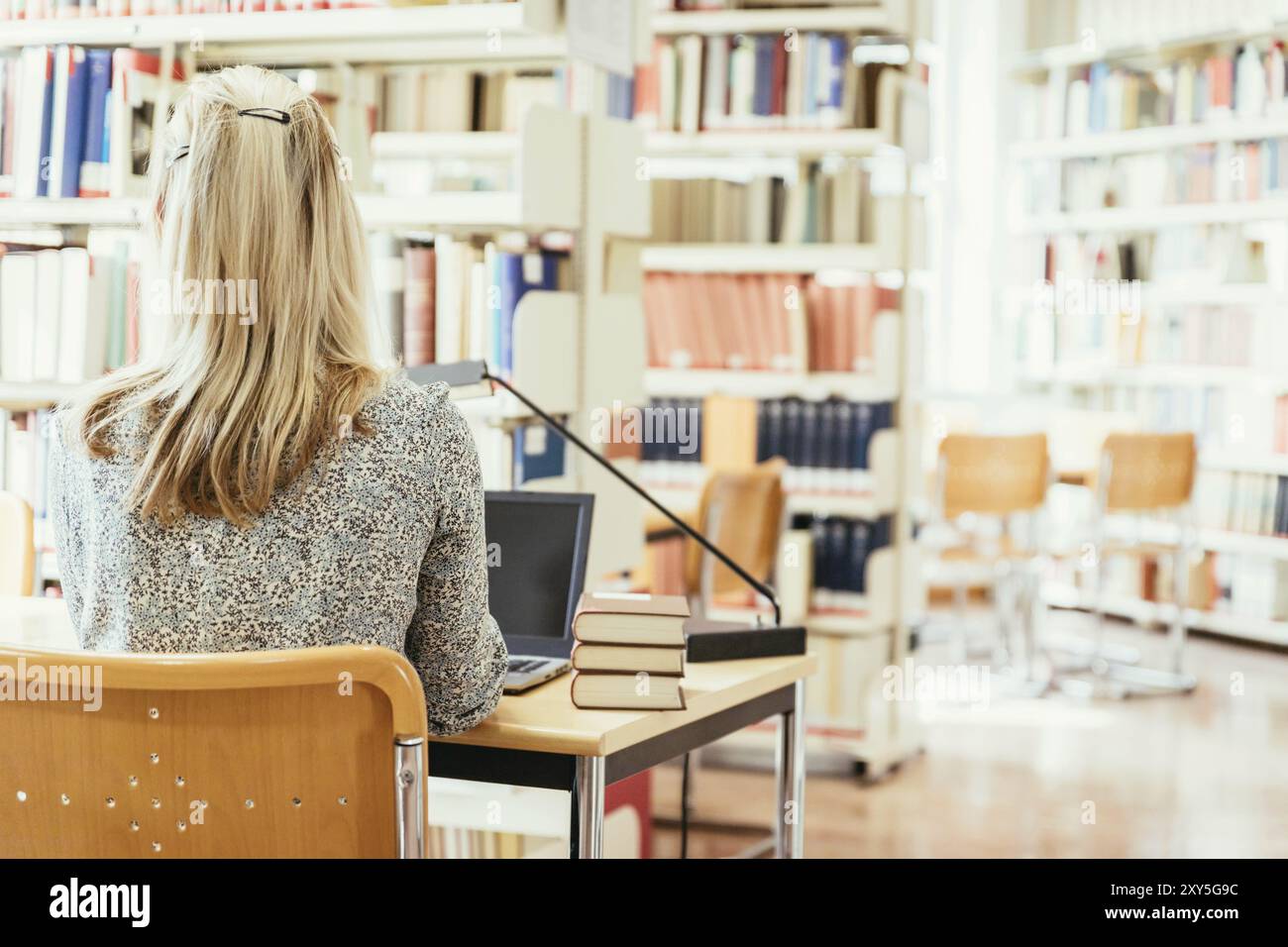 Blonde female student is sitting at the desk with pile of books ...