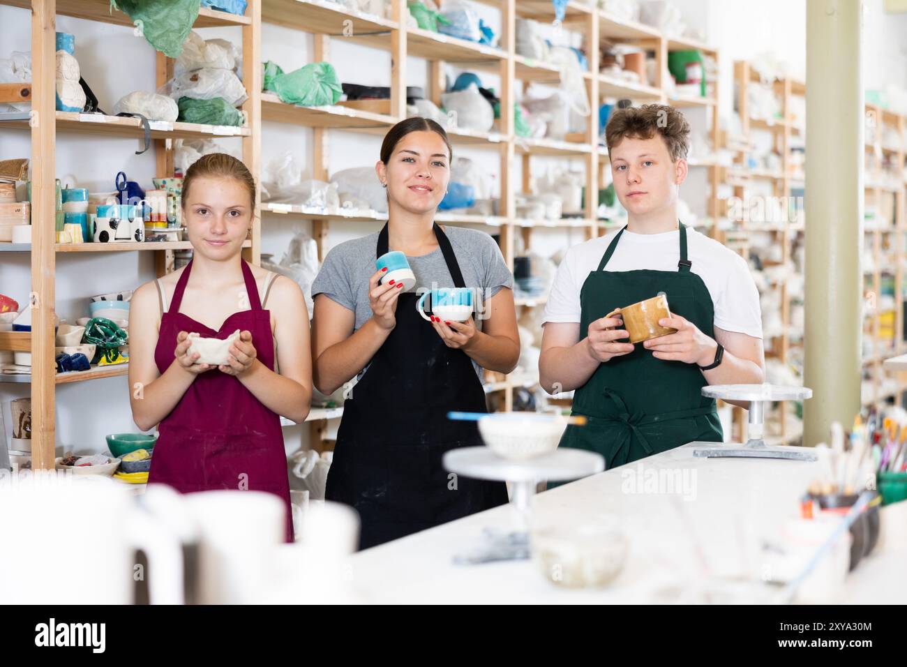 Young students, together with pottery teacher, show clay mugs they made ...
