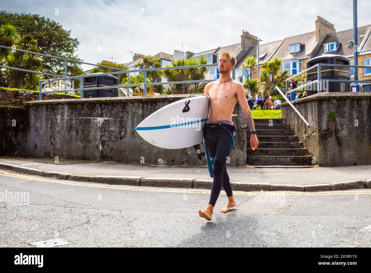 A surfer carrying his surfboard walking across a road in Newquay town ...