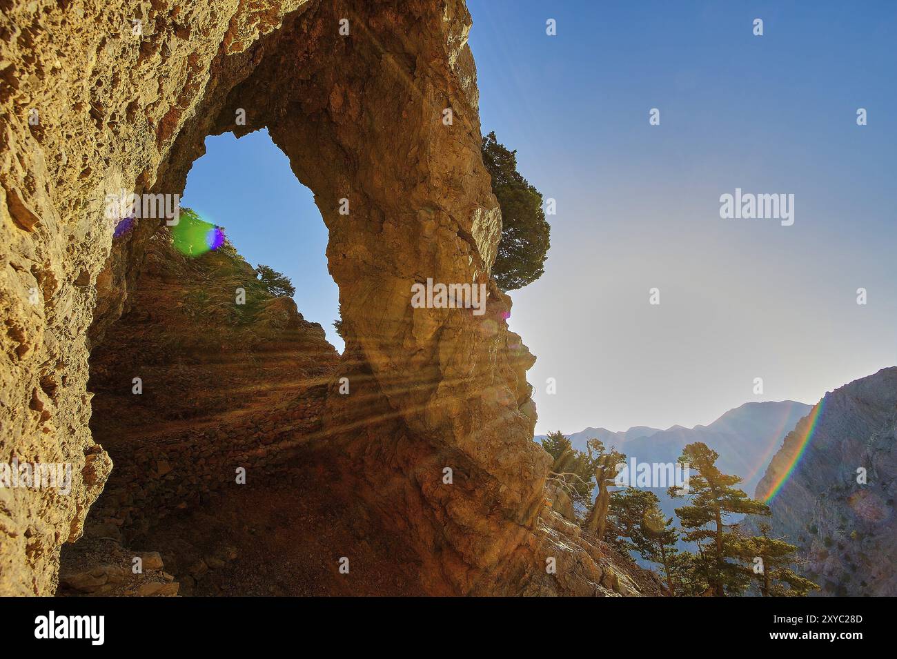 A rocky arch with sunbeams, surrounded by trees and mountains under a ...