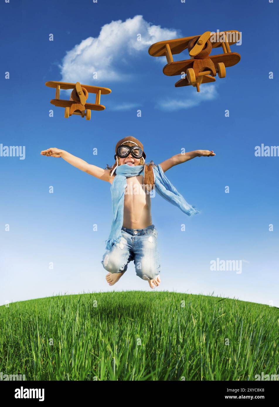 Happy kid dressed as a pilot jumping in green field against blue sky ...