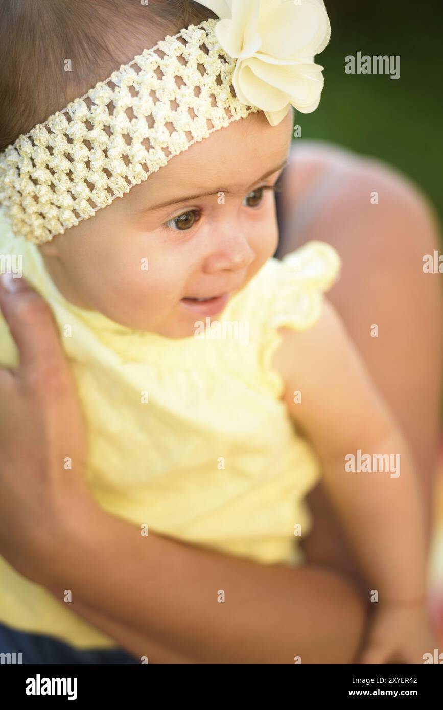 Portrait young mother holding cute baby girl in yellow band and dress. Sunny summer day outdoors ...