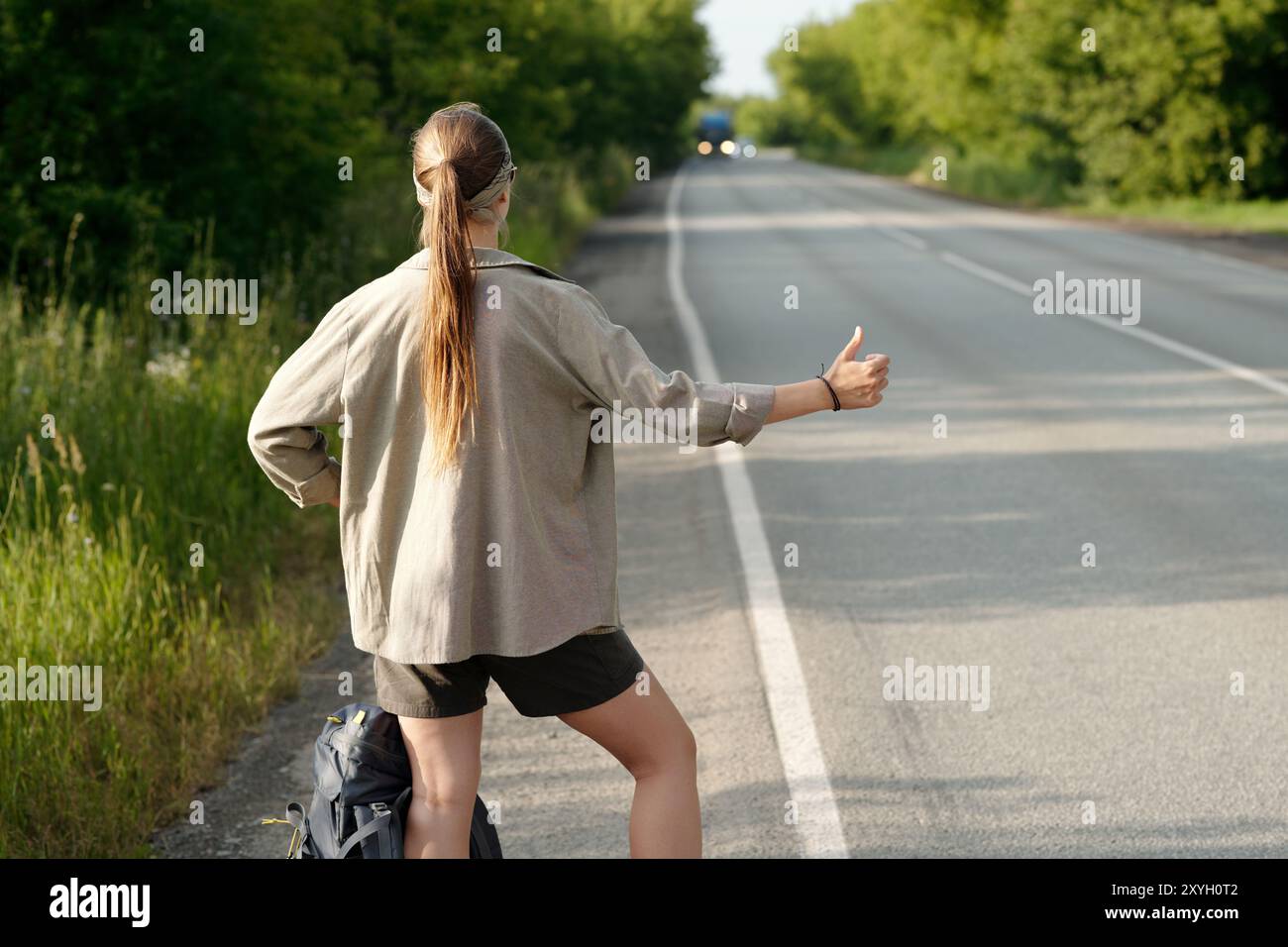 Back View Of Young Active Female Hitchhiker In Grey Shirt And Black Shorts Catching Car On