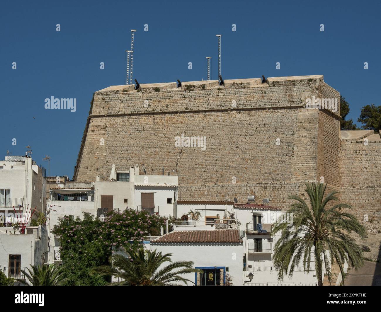 Historic stone walls above modern buildings and palm trees under a blue ...