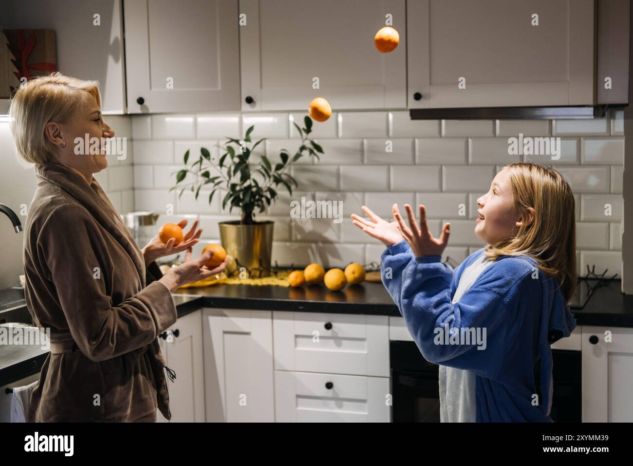 Mother and daughter juggling oranges in kitchen, playful family bonding ...