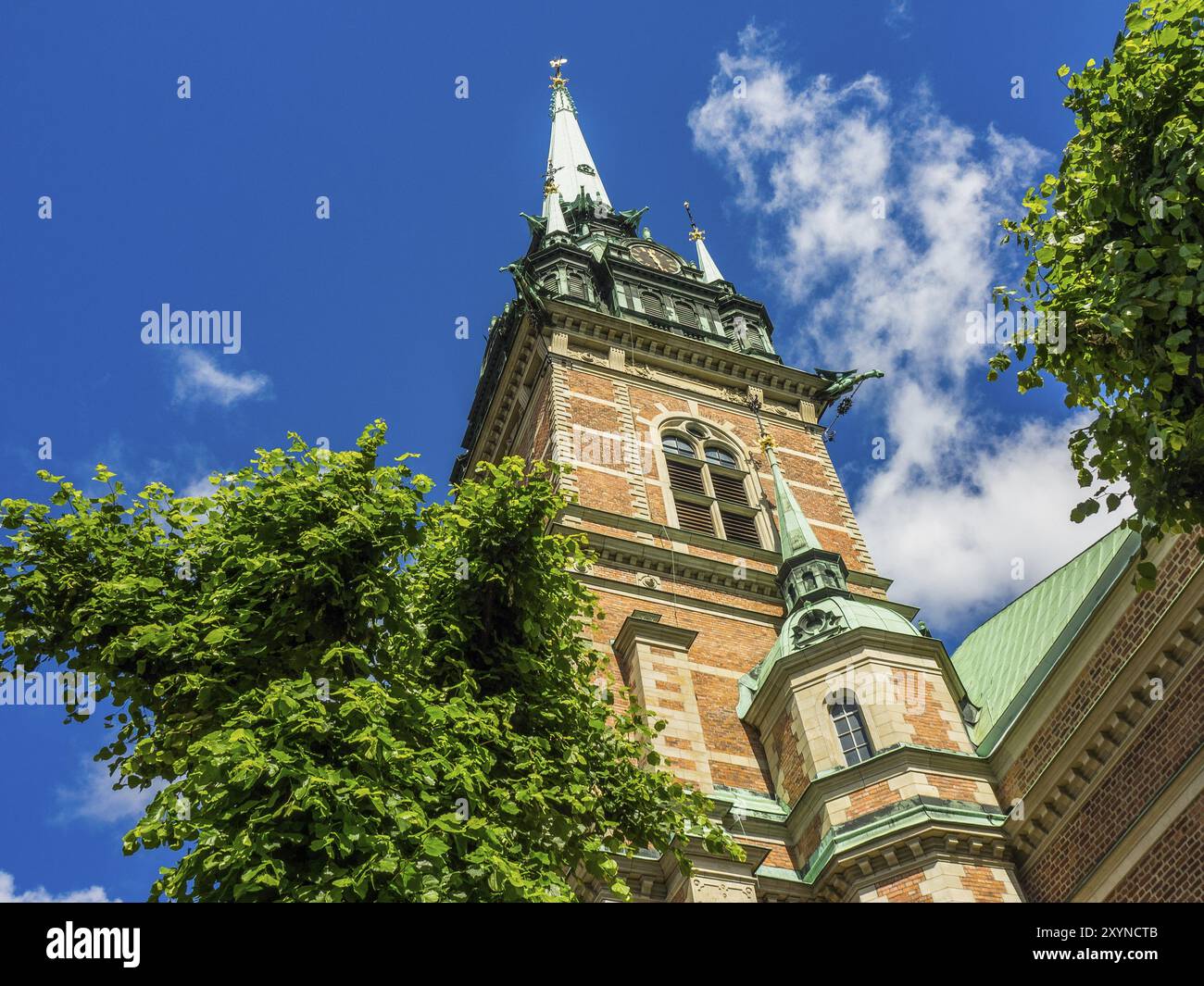 Gothic brick church tower flanked by green trees and a bright blue sky ...