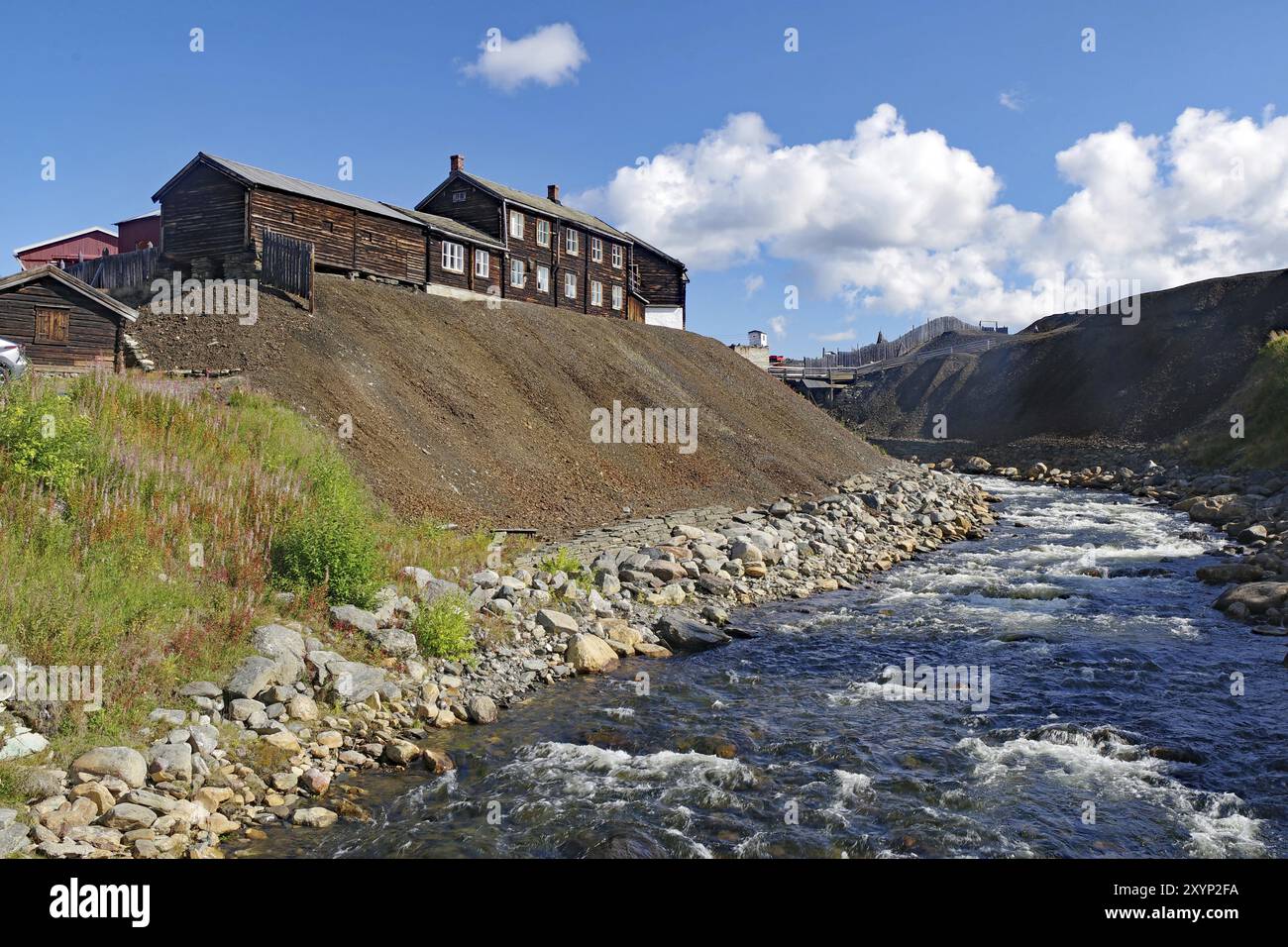A rushing river next to old buildings on a hill under a sky with white ...