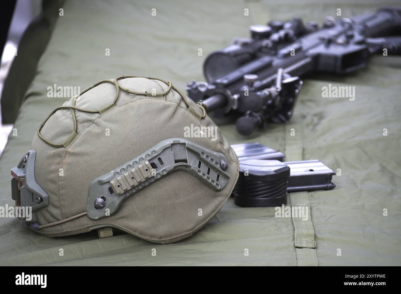 Military helmet alongside rifle with a scope on a cloth surface, also ...