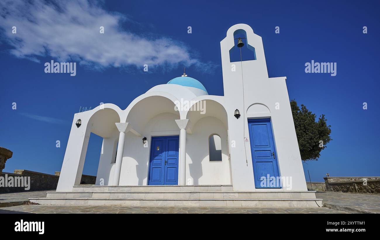 White church with blue doors and a bell in front of a blue sky and tree ...