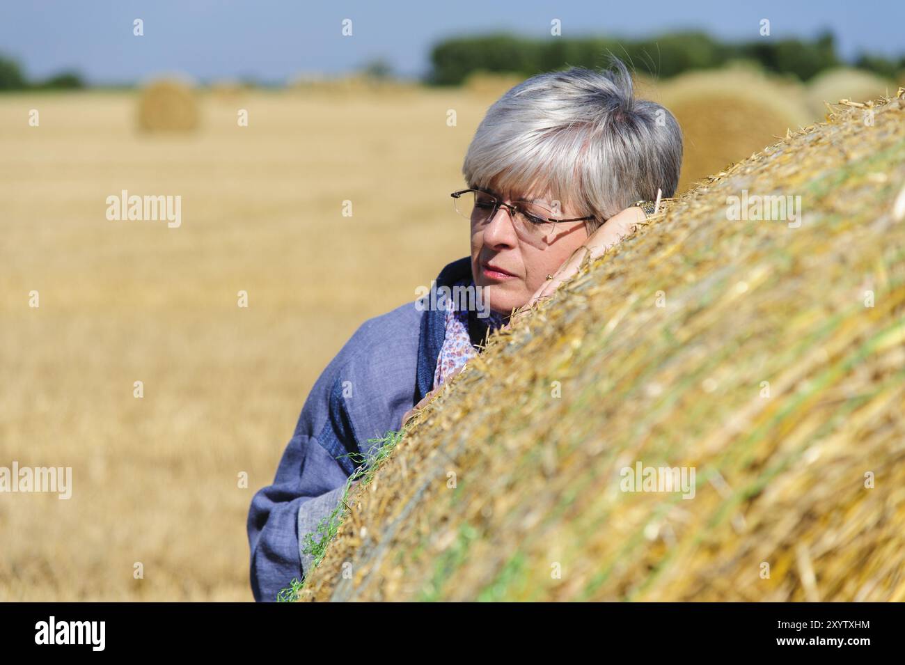 Grey-haired lady leans her head against a bale of straw and rests Stock ...