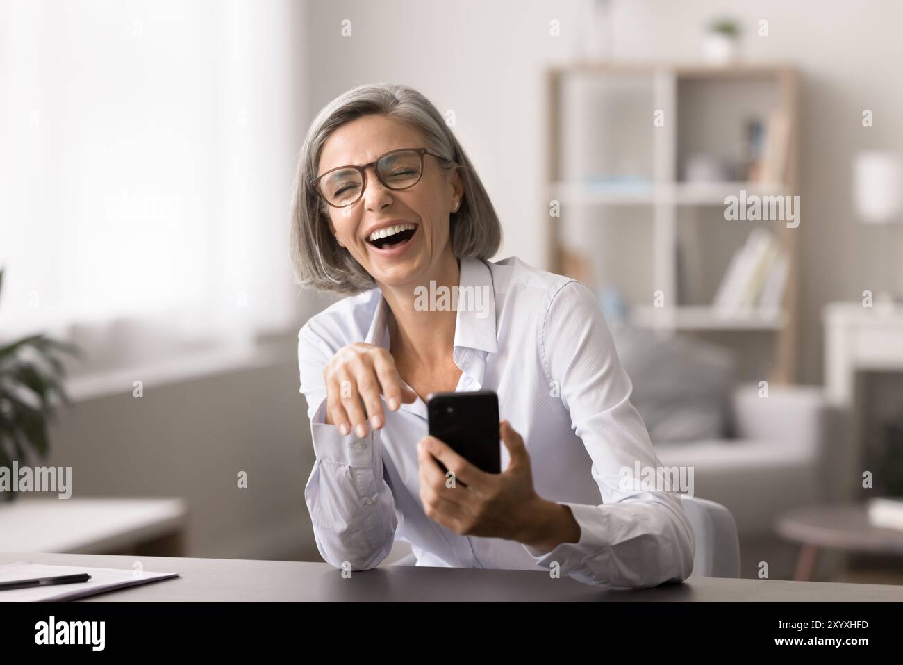 Happy middle-aged woman laughing seated at desk with modern smartphone Stock Photo - Alamy