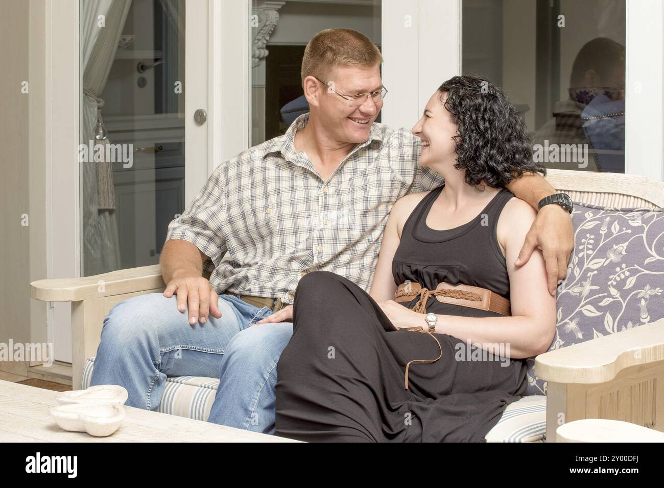 A young couple sit togehter on a bench out on the patio, man has arm ...