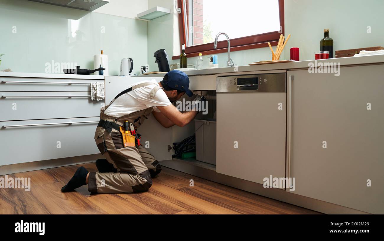 Young professional male plumber in uniform is repairing a kitchen sink ...