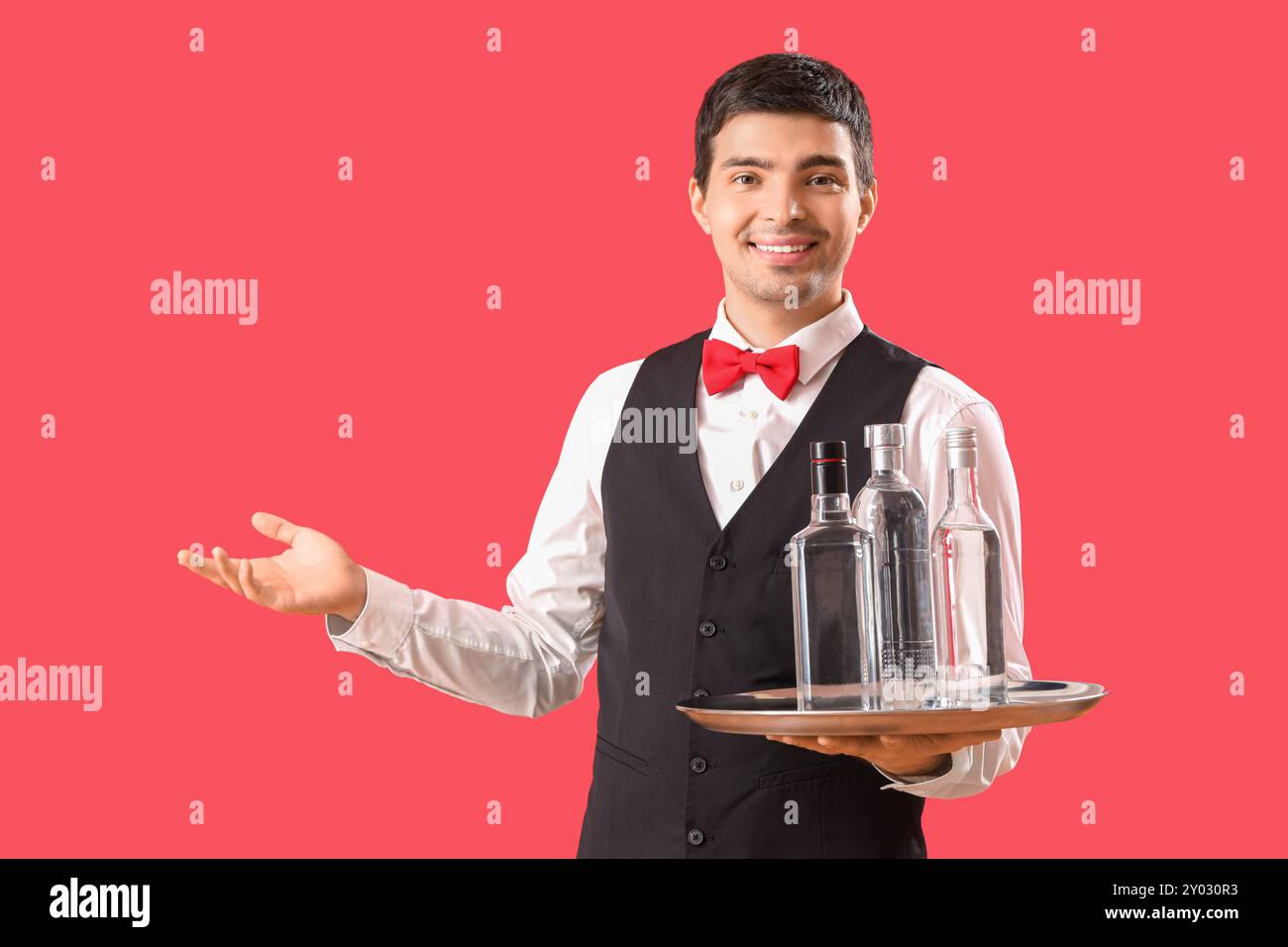 Male waiter with bottles of vodka showing something on red background ...