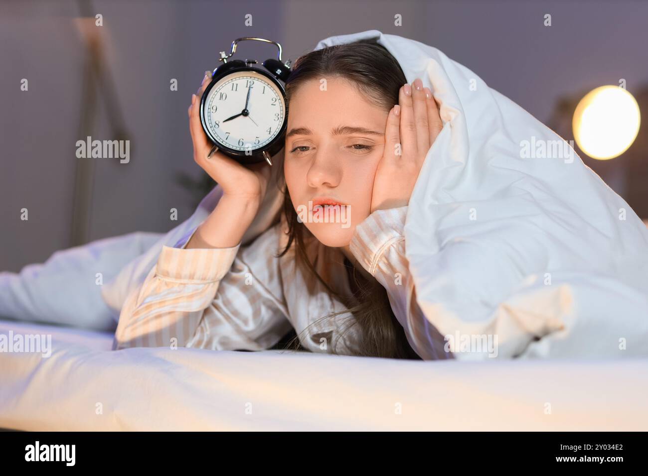 Sleepy young woman with alarm clock lying under blanket in bedroom at ...
