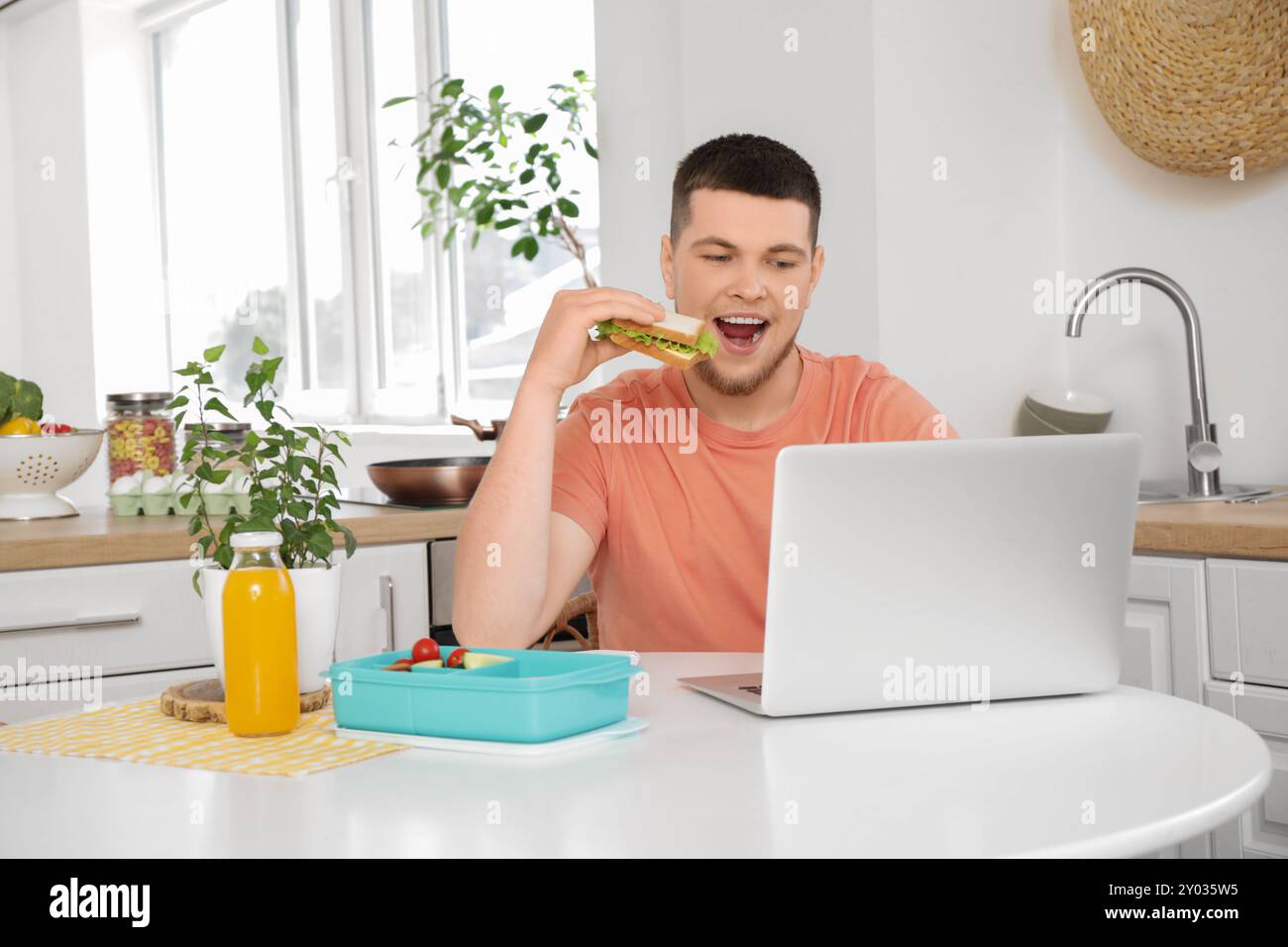 Young man with laptop and lunch box eating tasty sandwich in kitchen ...