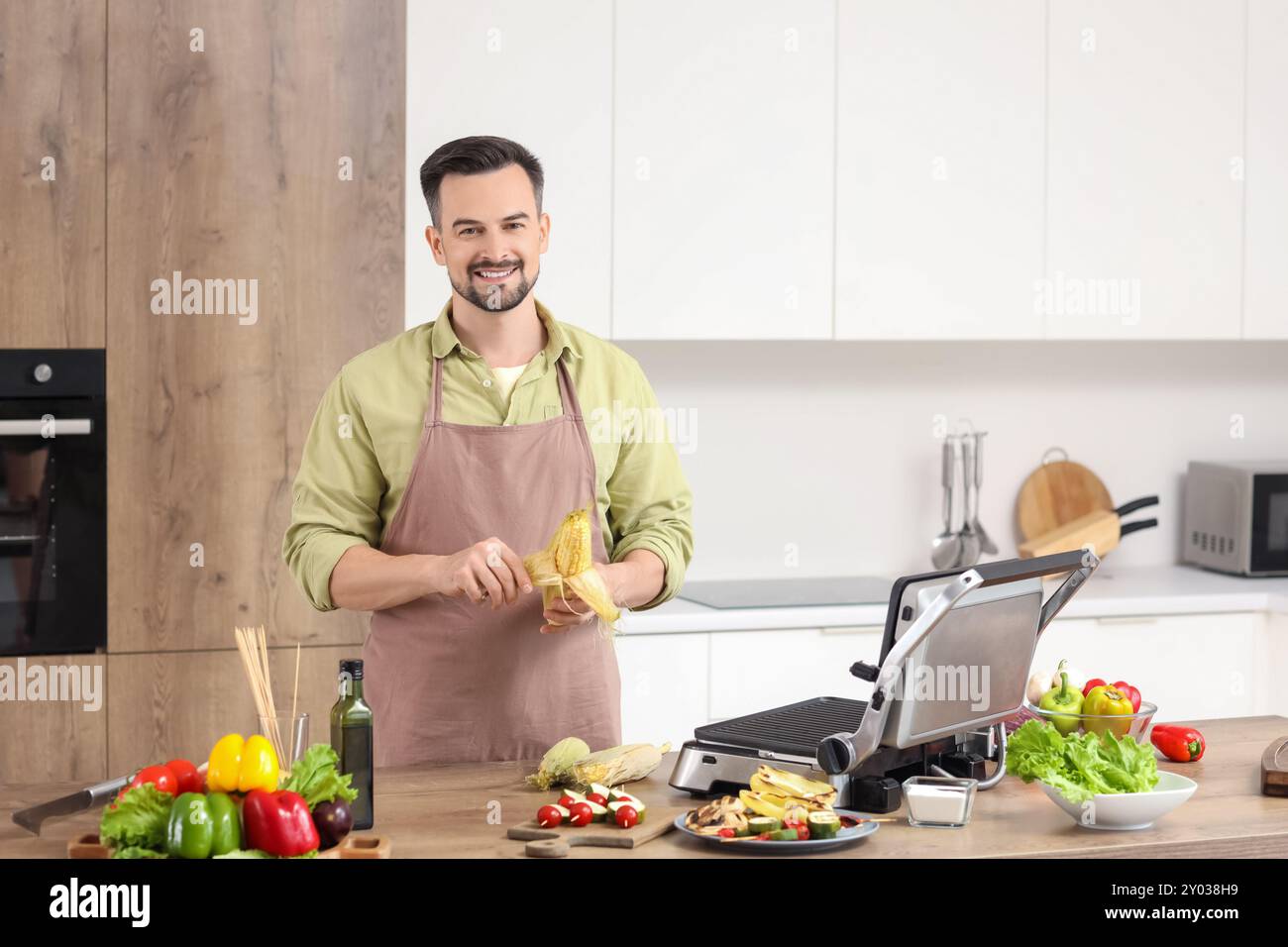 Young happy man peeling corn cobs to cooking on modern electric grill ...