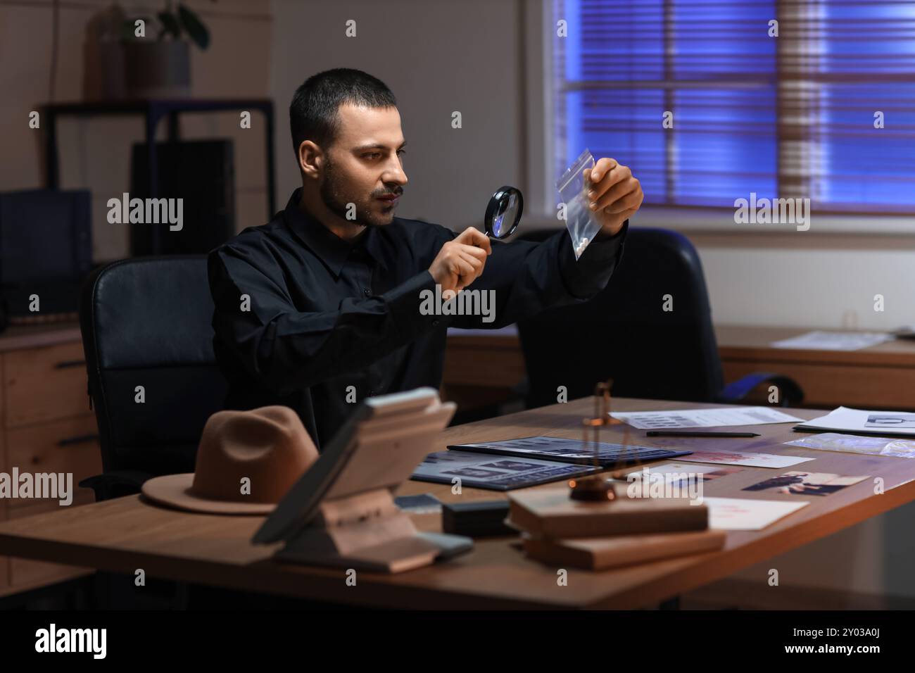Male detective with magnifier examining evidence in office at night ...
