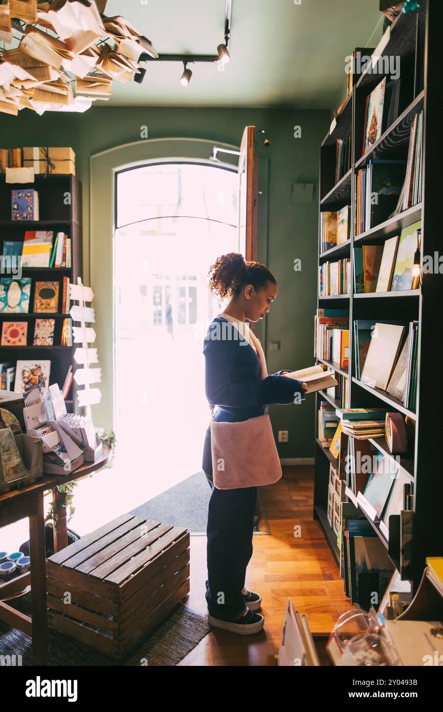 Side view of woman reading book while standing near bookshelf at store ...