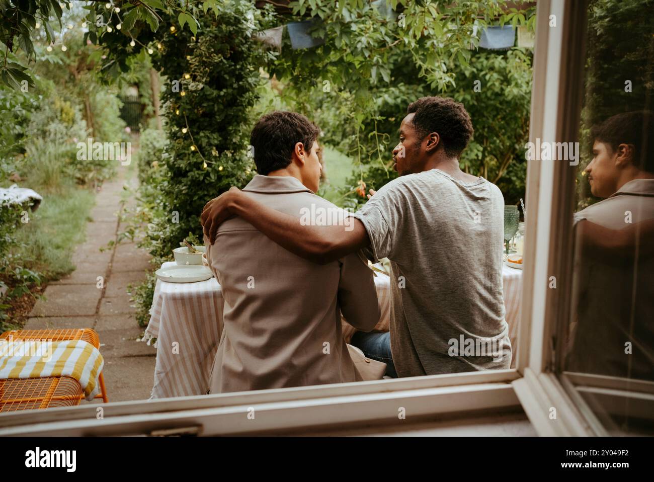 Rear view of young man sitting with arm around male friend near window ...