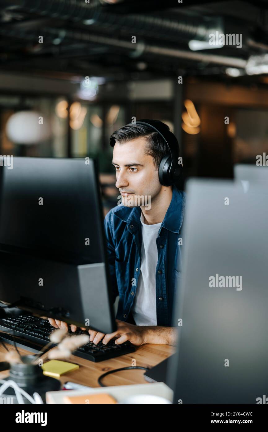 Young computer programmer typing on desktop PC with wireless headphones ...
