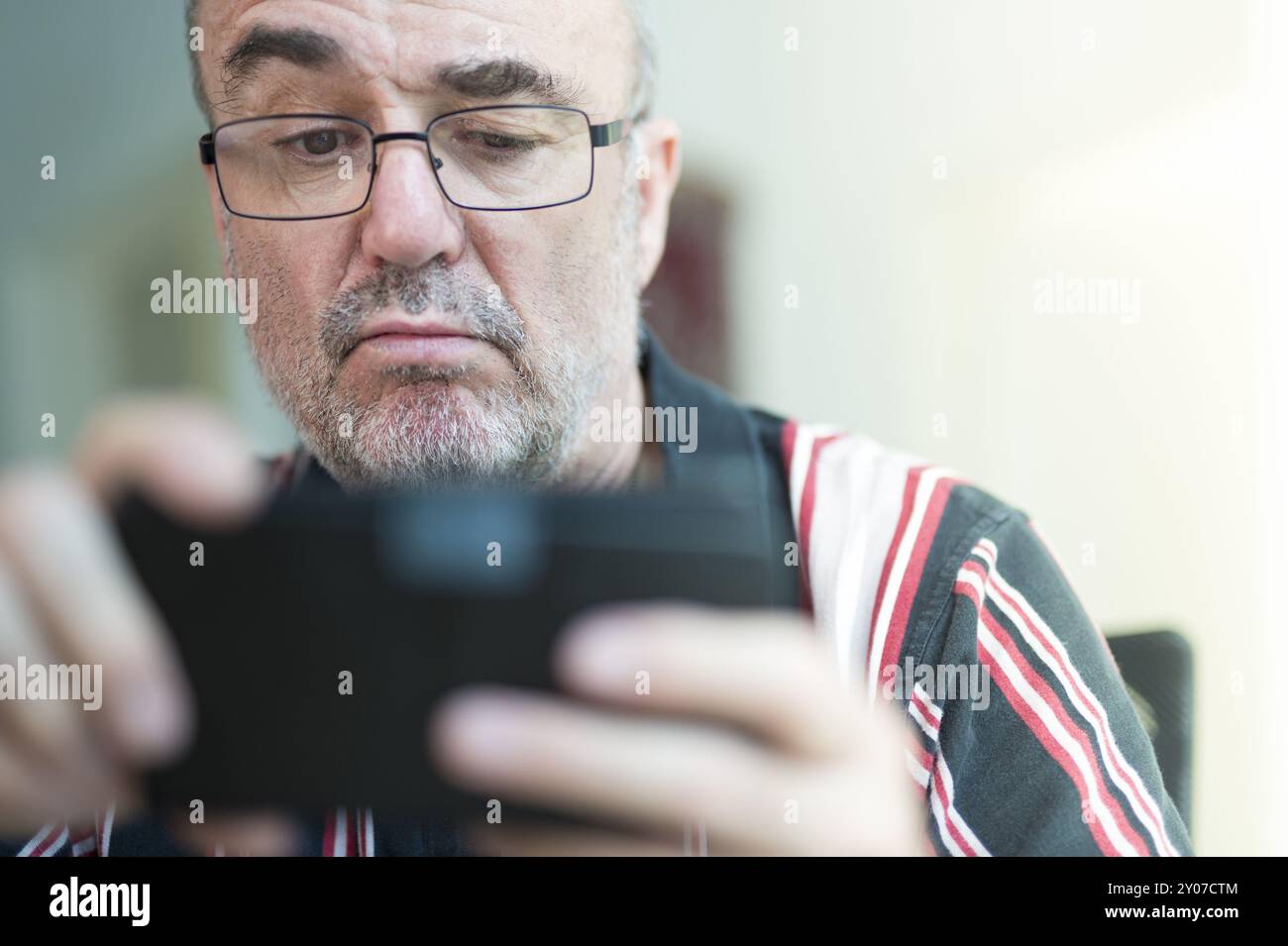 An old man holds a smartphone in his hands and looks sceptically at the ...