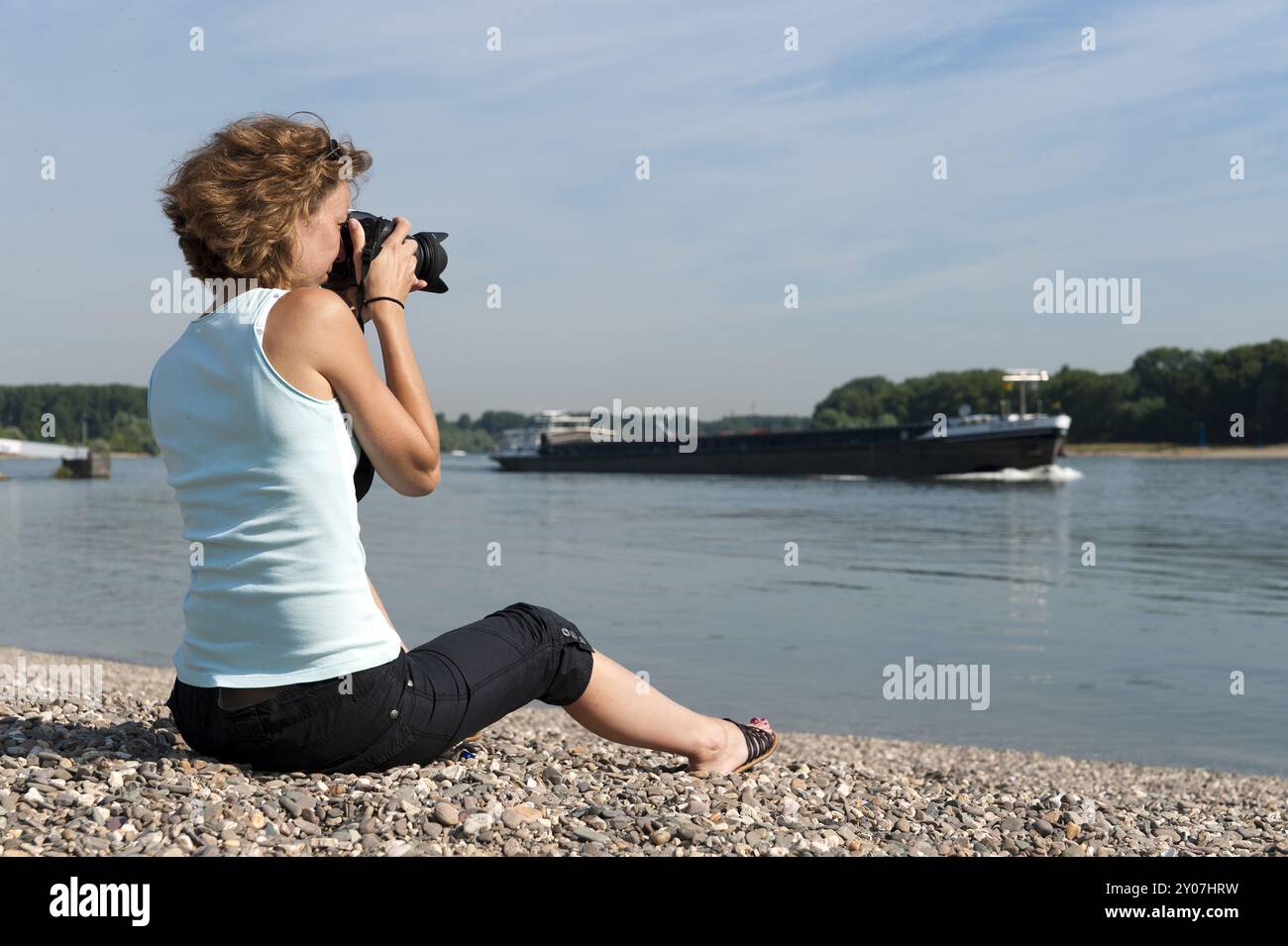Young woman sits on the Rhine and photographs a barge Stock Photo - Alamy
